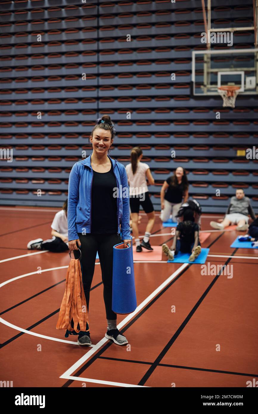 Full length portrait of smiling female coach standing with exercise mat ...