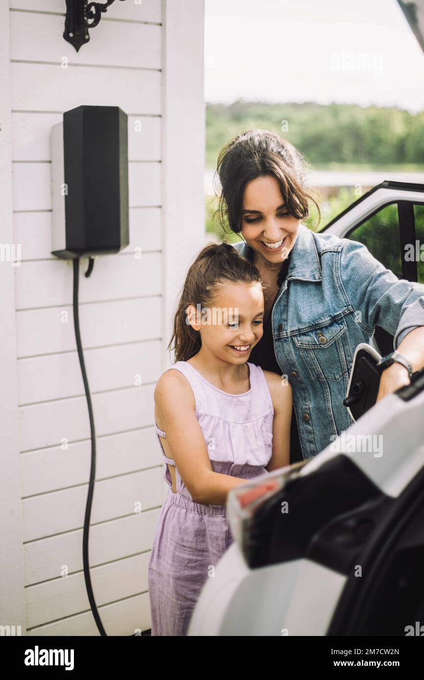 Happy mother and daughter charging electric car together near house ...
