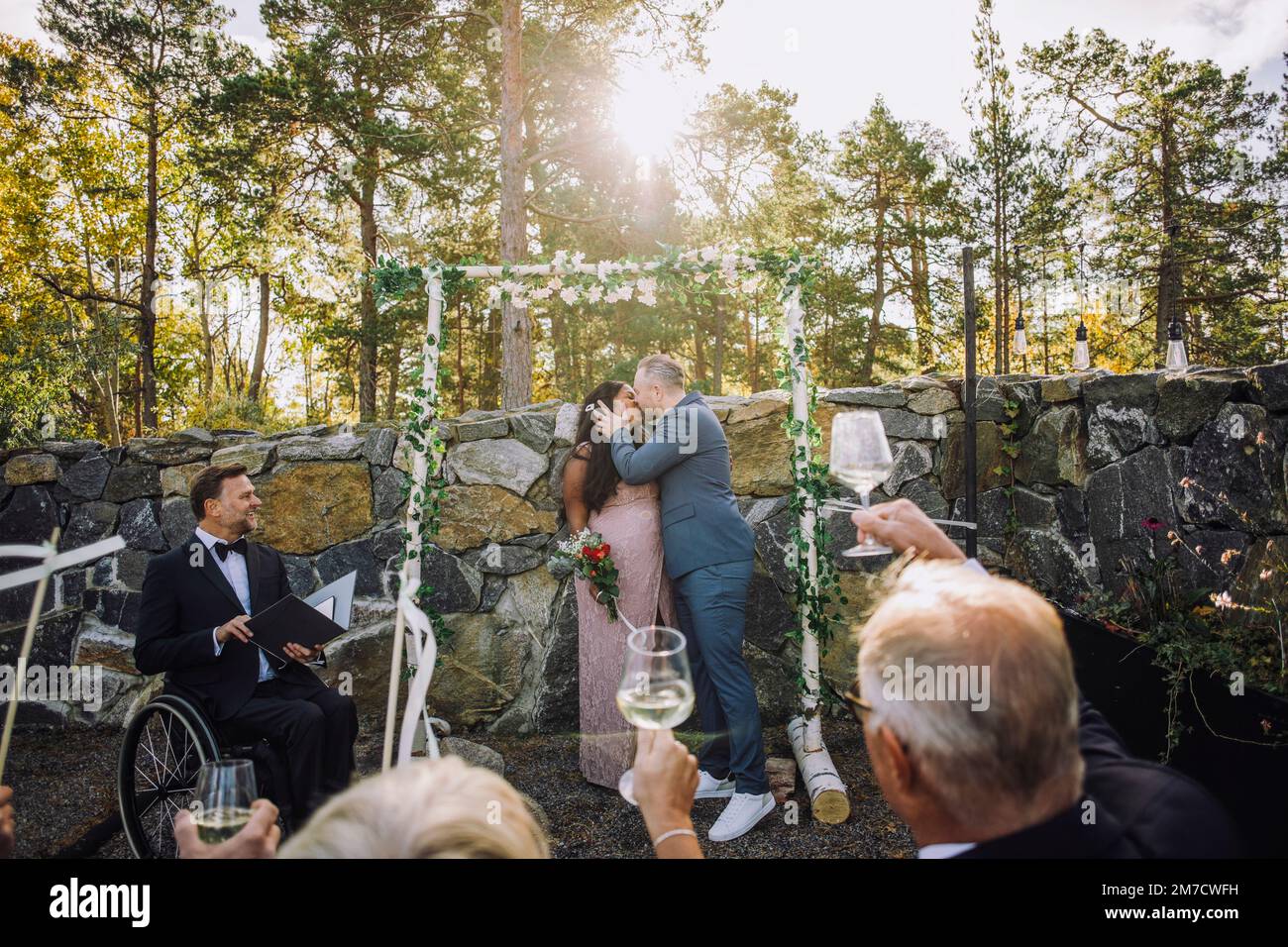 Newlywed couple kissing in front of guests raising toasts and minister ...