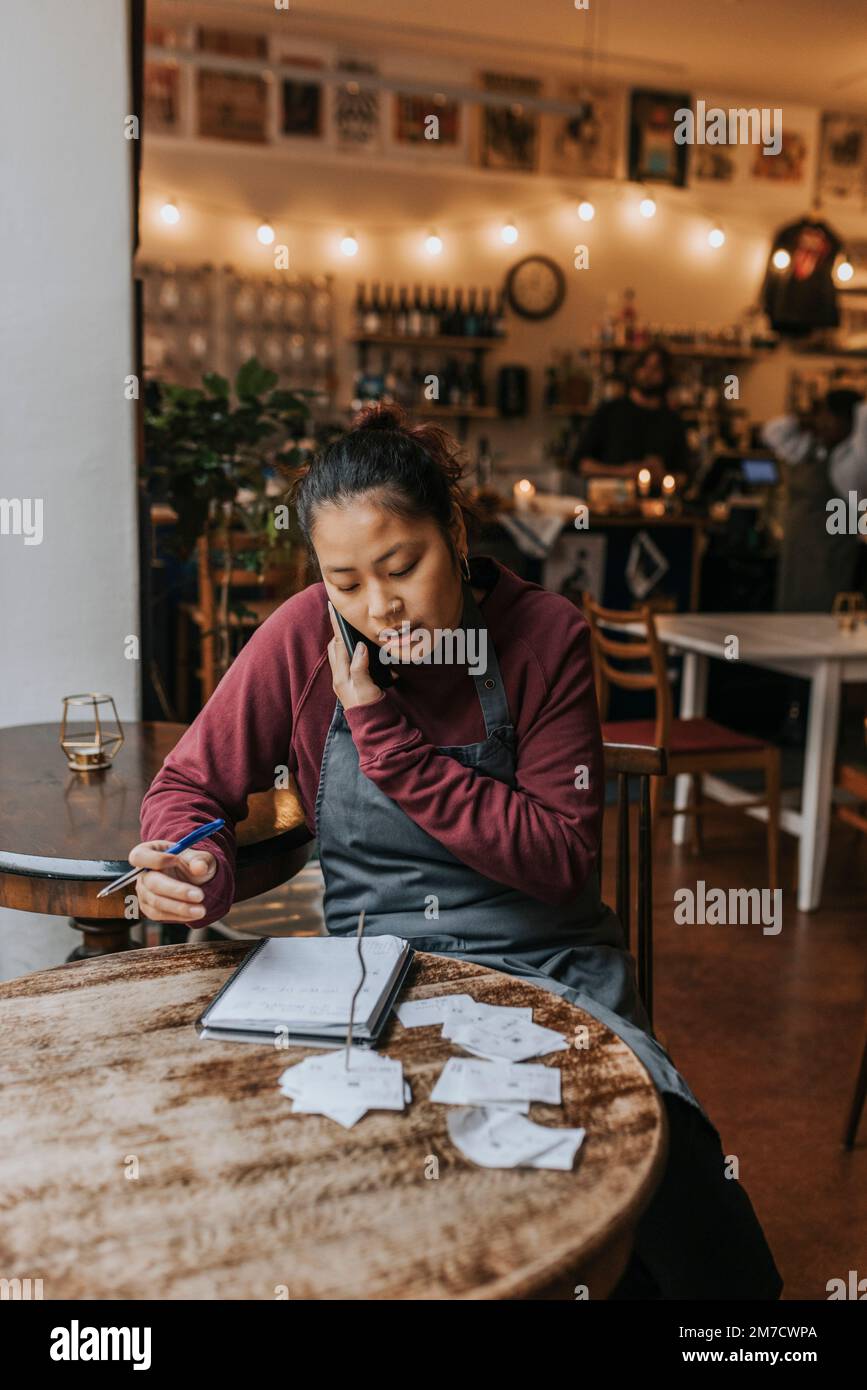 Female cafe owner talking on mobile phone while sitting with diary at ...