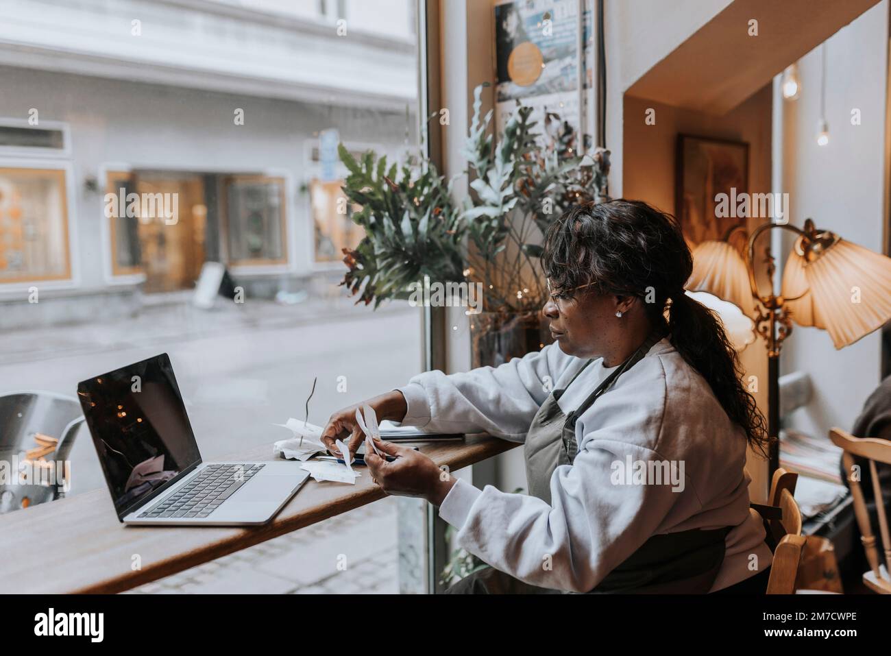 Side view of mature cafe owner with bills and laptop sitting at table ...