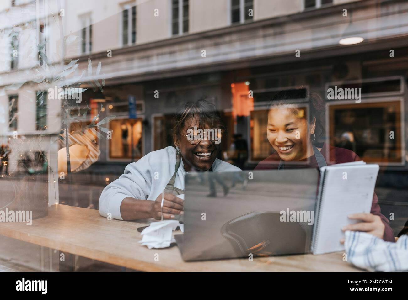Happy female coffee shop owners discussing over laptop seen through ...