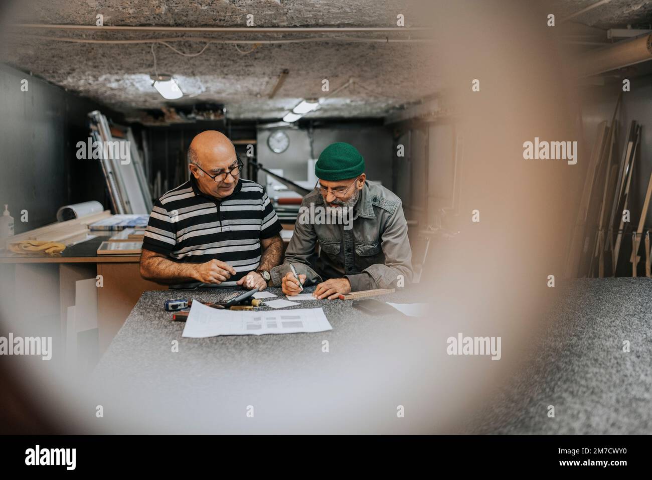 Male craftsman writing while discussing with colleague at workbench ...