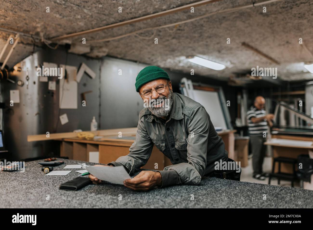 Portrait of cheerful bearded craftsman with blueprint leaning on ...