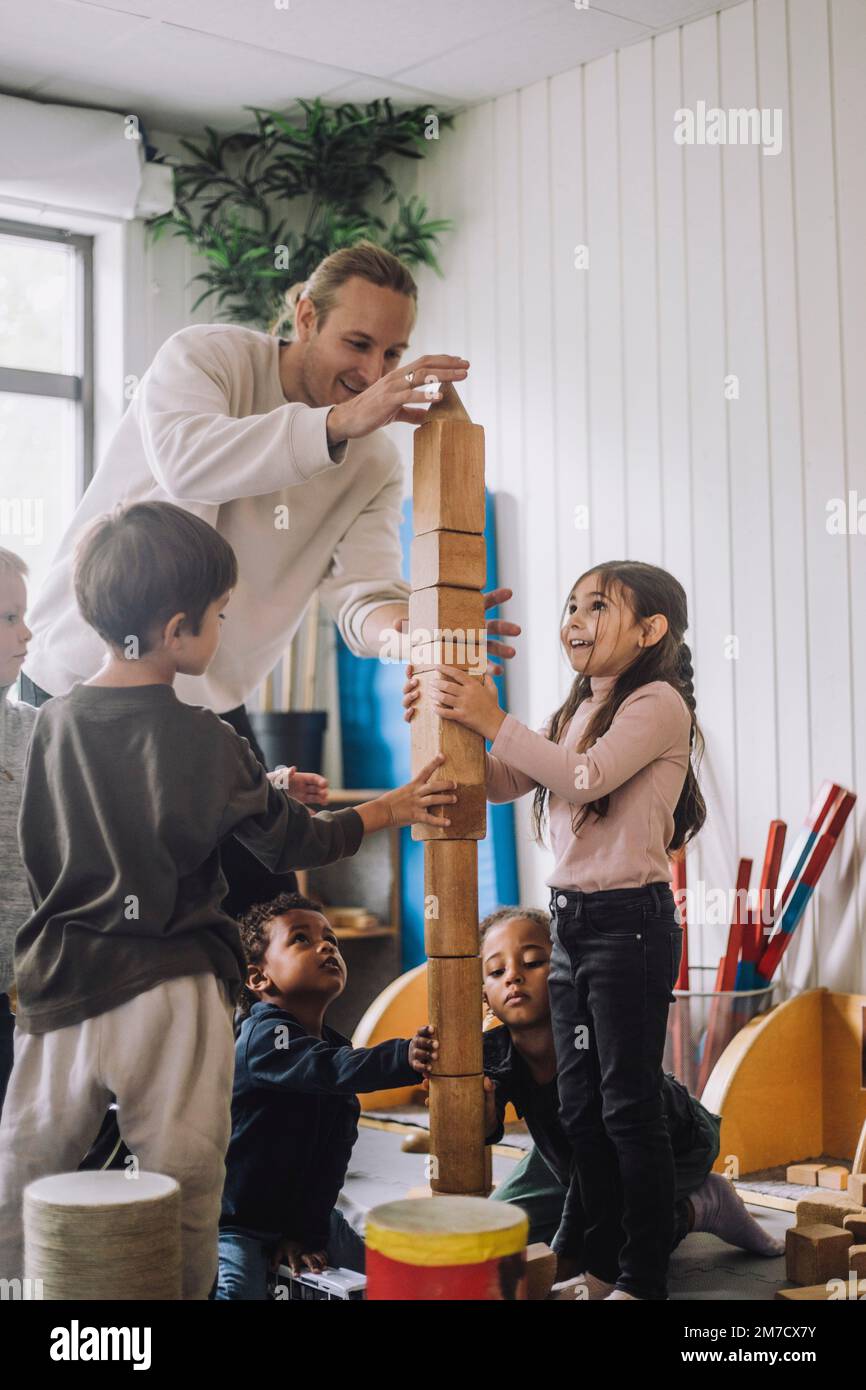 Male teacher guiding children stacking wooden toy blocks at ...