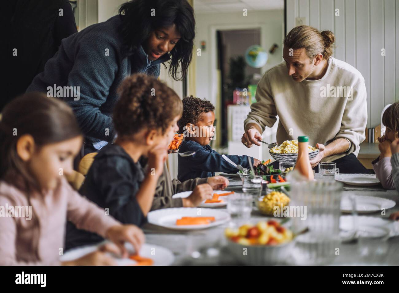 Male and female teacher serving food to children while having meal at ...
