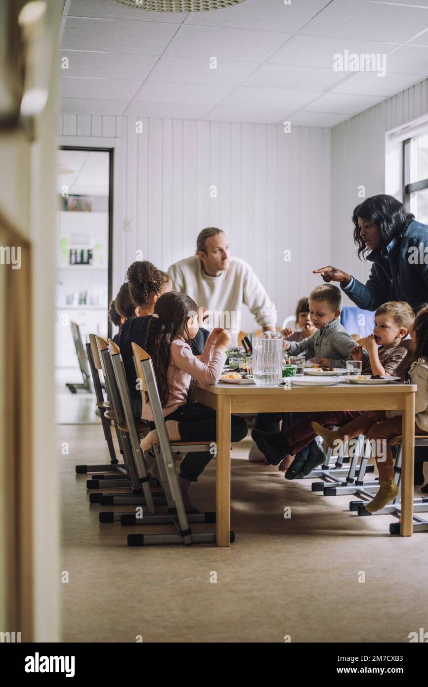 Female and male teachers talking with children having breakfast at ...