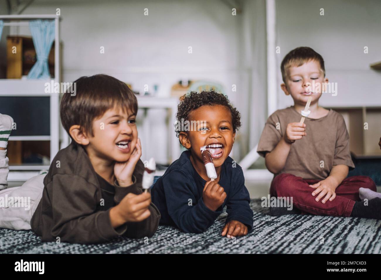 Happy multiracial male students eating ice creams on carpet at day care ...