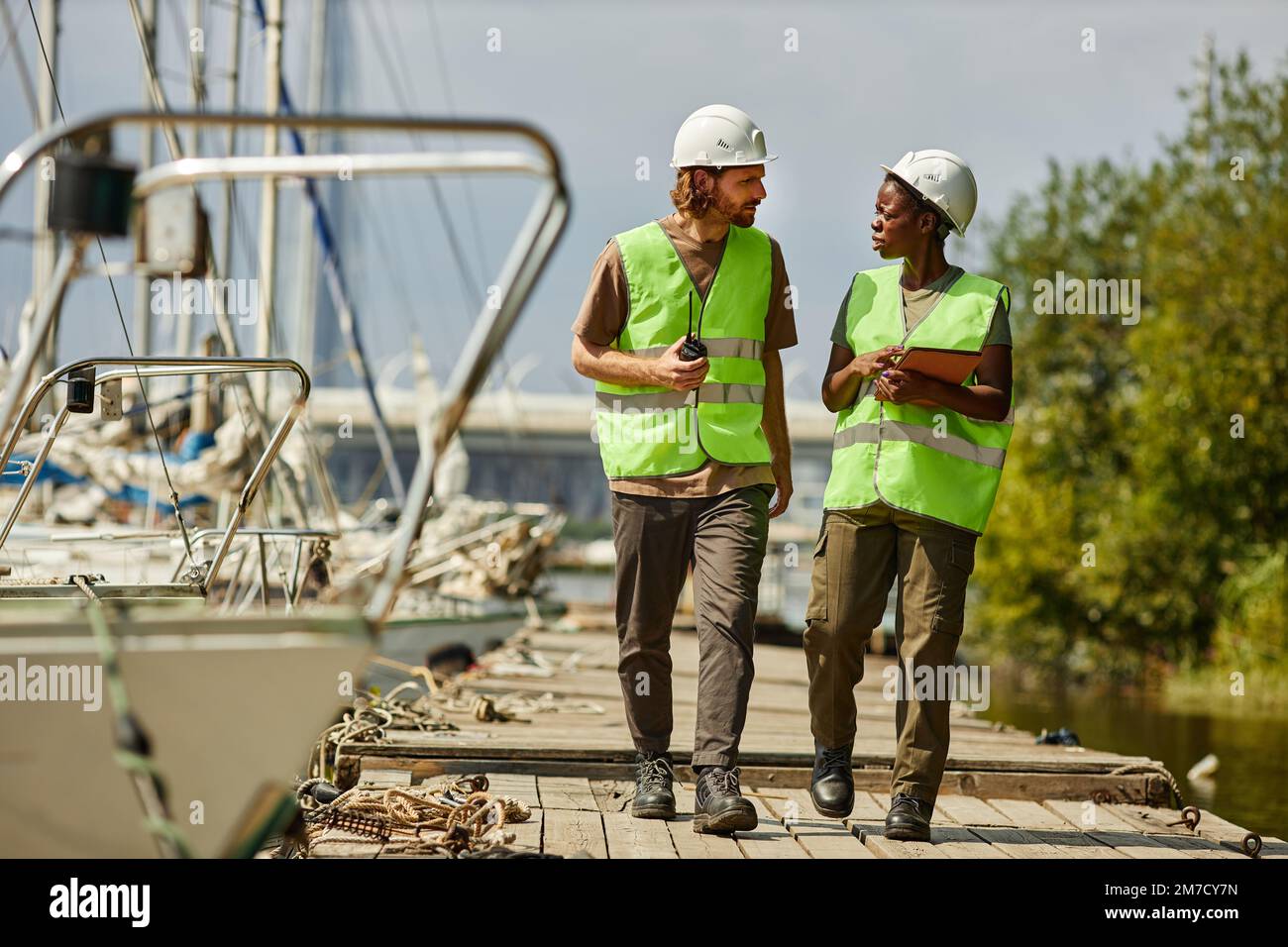 Full length portrait of two young workers wearing hardhats while walking towards camera on pier ...
