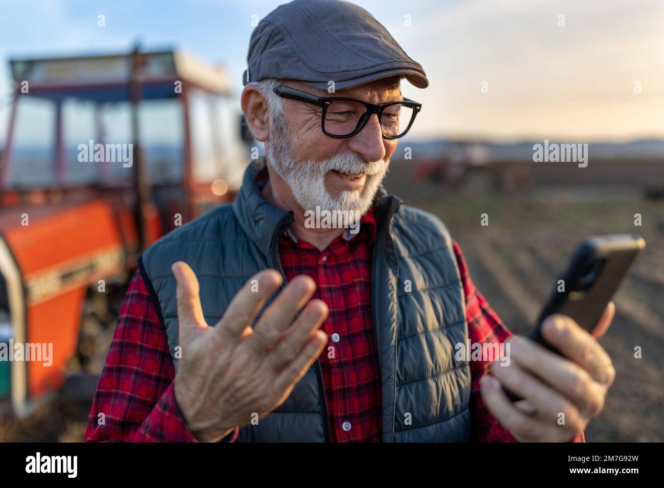 Excited mature farmer talking on mobile phone in front of tractor in ...