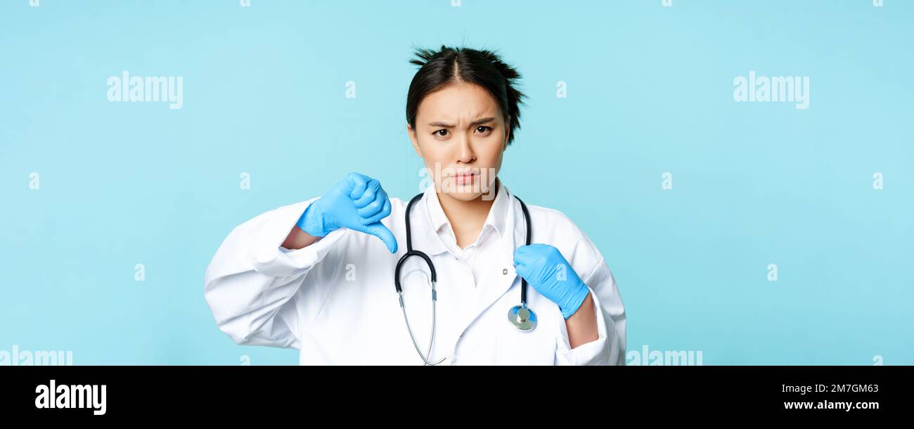Angry asian woman doctor, physician in uniform and gloves shows thumbs ...