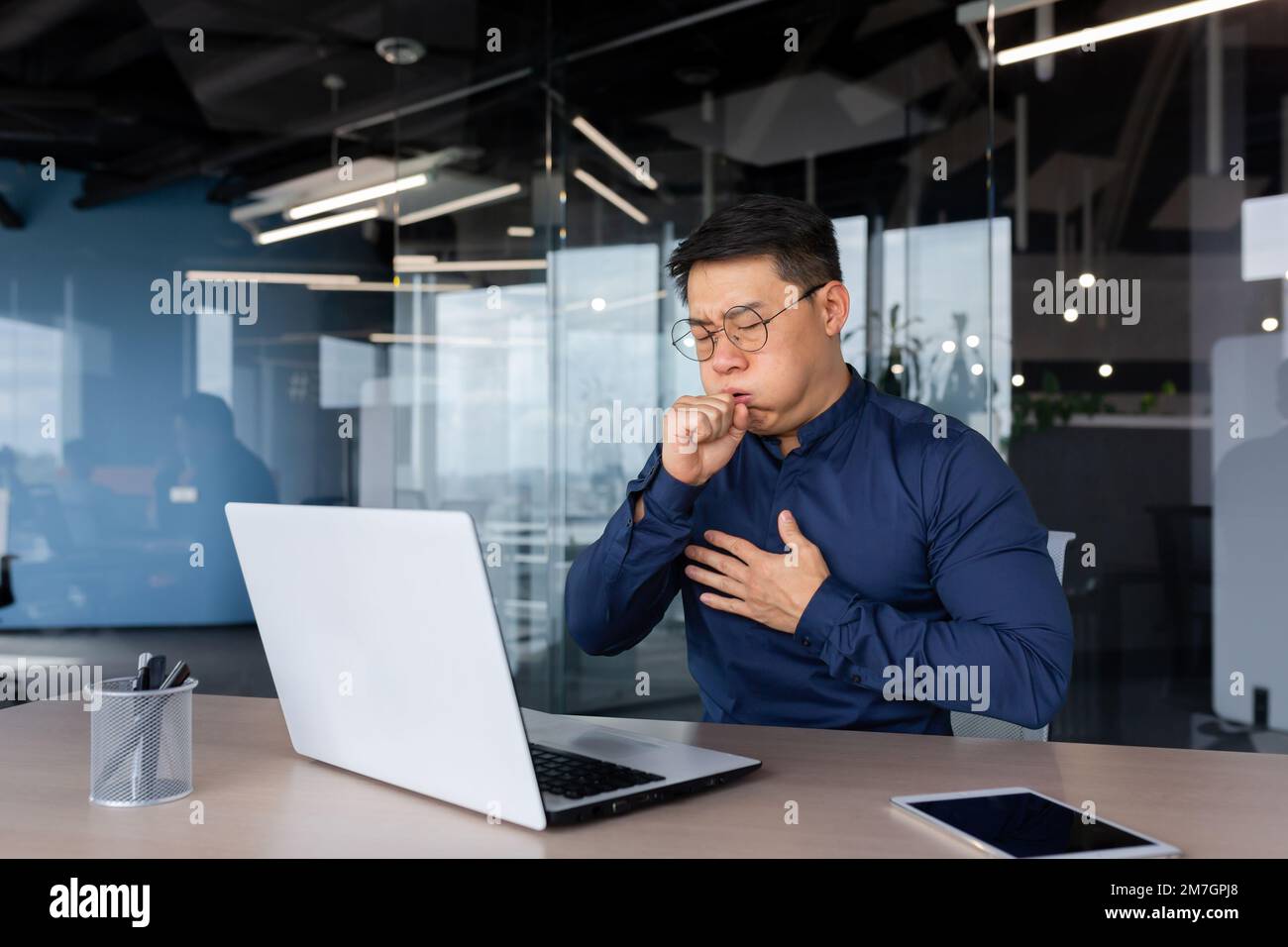 A young Asian man coughs in the office at the desk. Has allergies, flu, asthma. He holds his ...