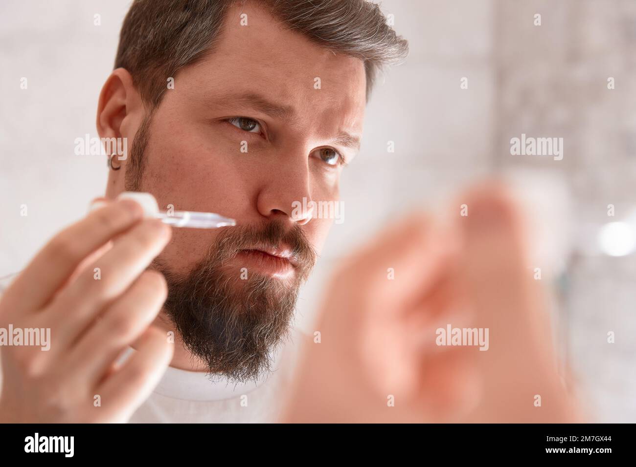 Portrait of man in white tshirt applying serum on his face at bathroom ...