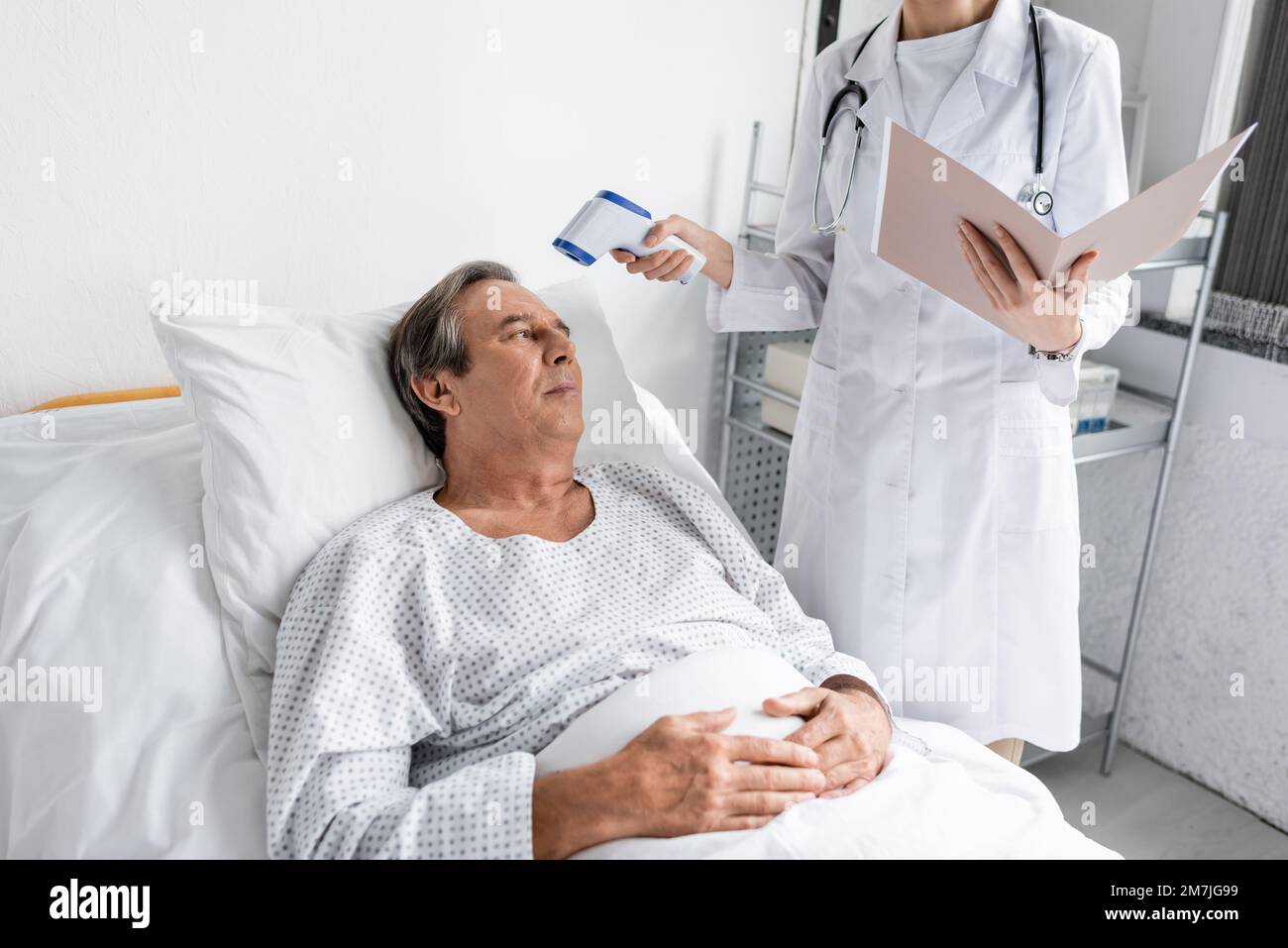 Doctor with paper folder holding pyrometer near senior patient in ...