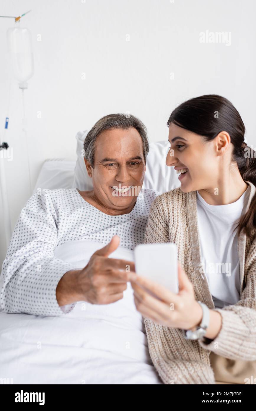 Positive patient pointing at smartphone near daughter in hospital ward ...