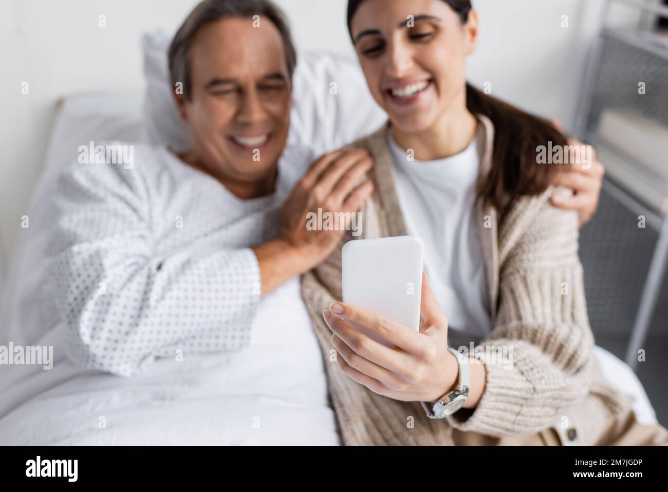 Blurred woman holding smartphone near elderly father on bed in hospital ...