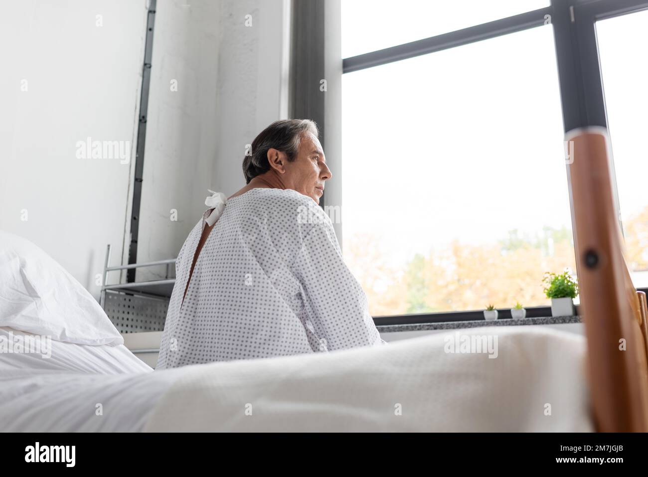 Side view of sick elderly man in patient gown sitting on blurred ...