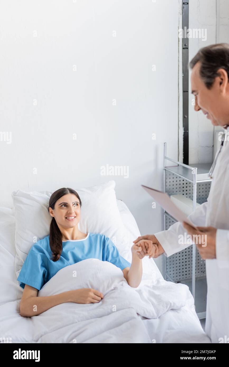 Smiling patient holding hand of elderly doctor with paper folder in ...