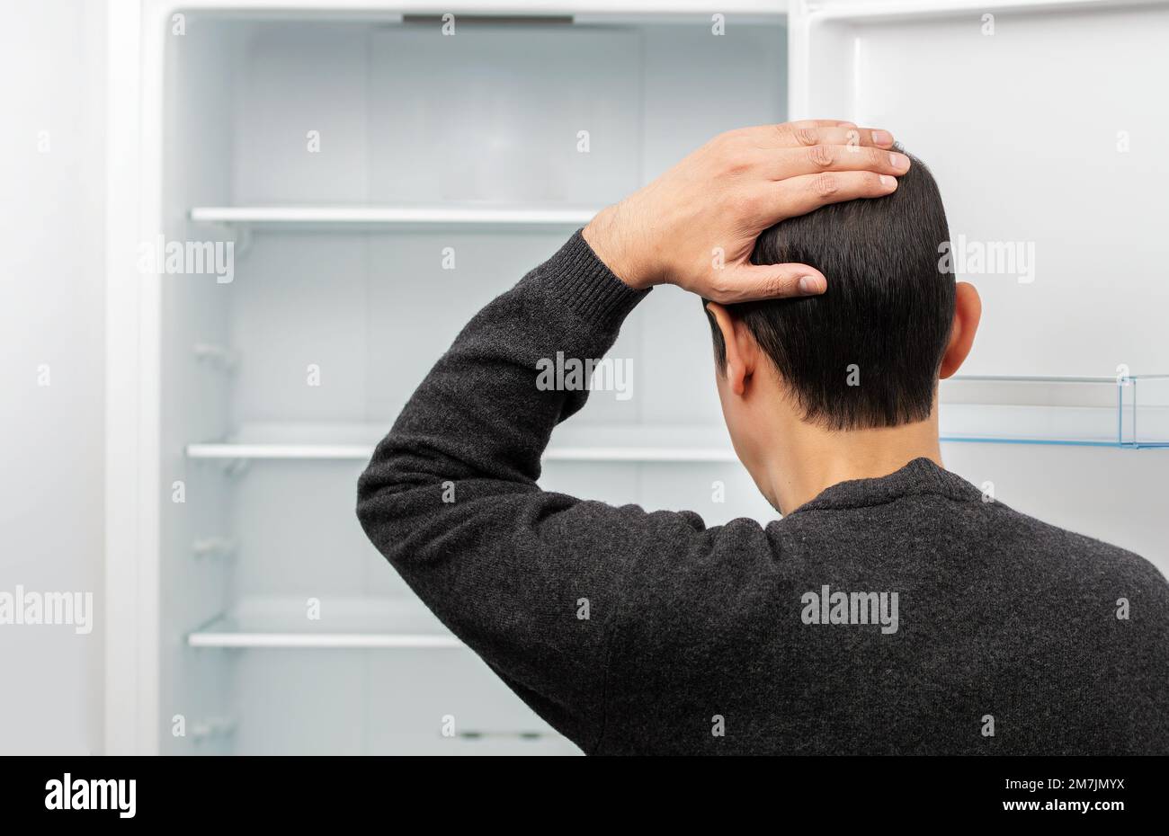 Rear view of a confused young man looking at food in refrigerator with ... Rear view of a confused young man looking at food in refrigerator with ...