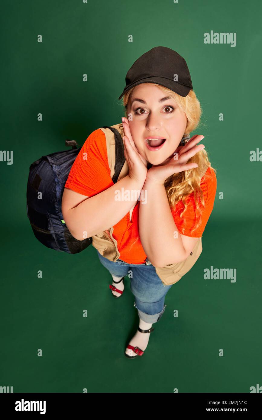 Portrait of blonde woman in cap with backpack posing over green studio ...
