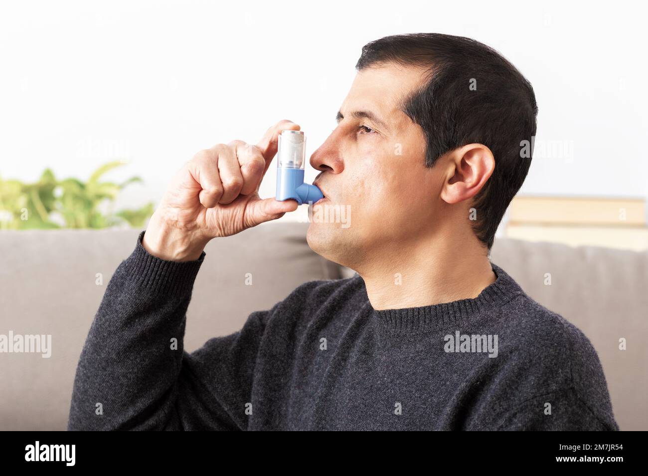 Side view of an asthmatic man using an inhaler sitting on a couch in ...