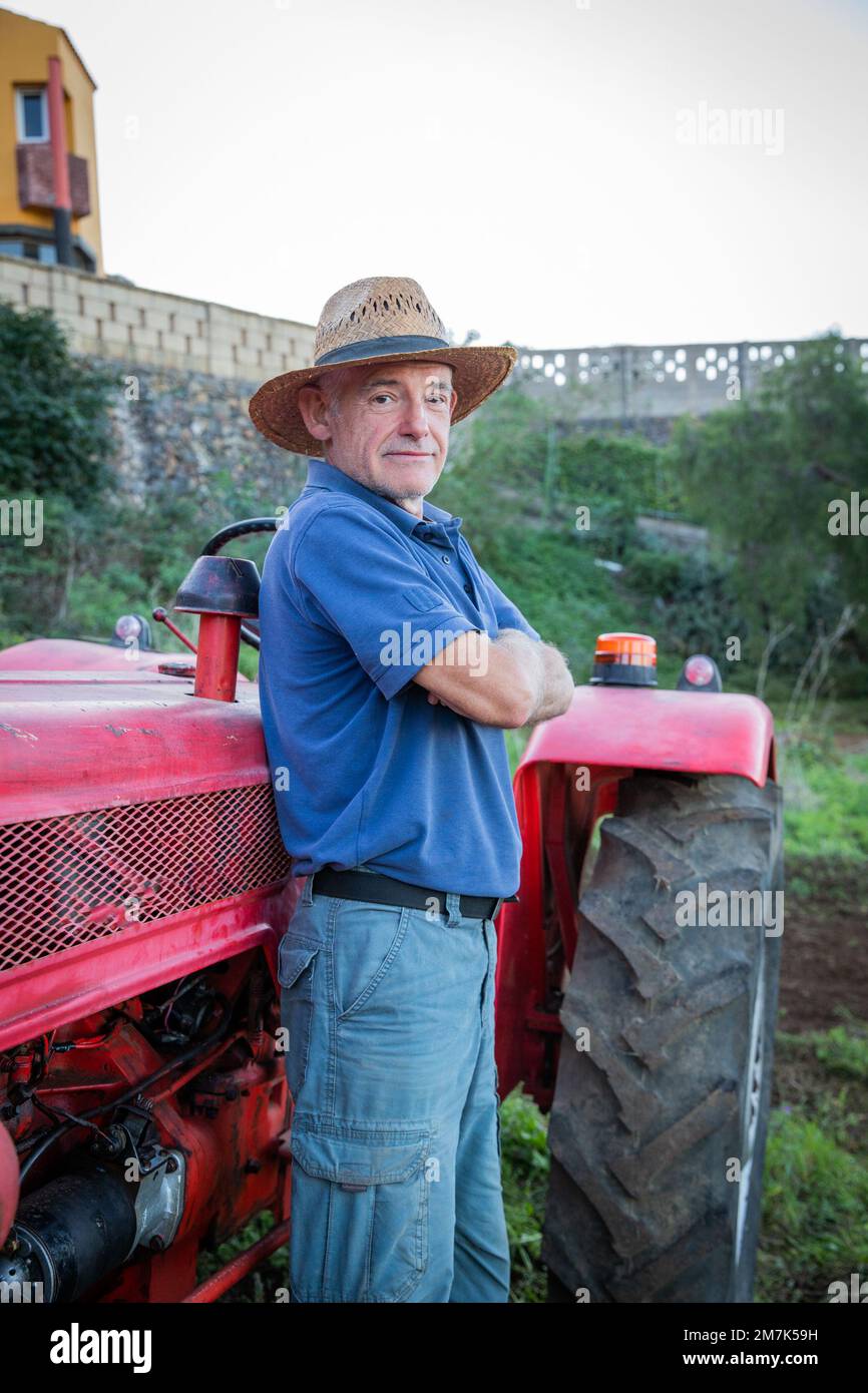 A farmer with his arms folded and his back against his tractor, mature ...