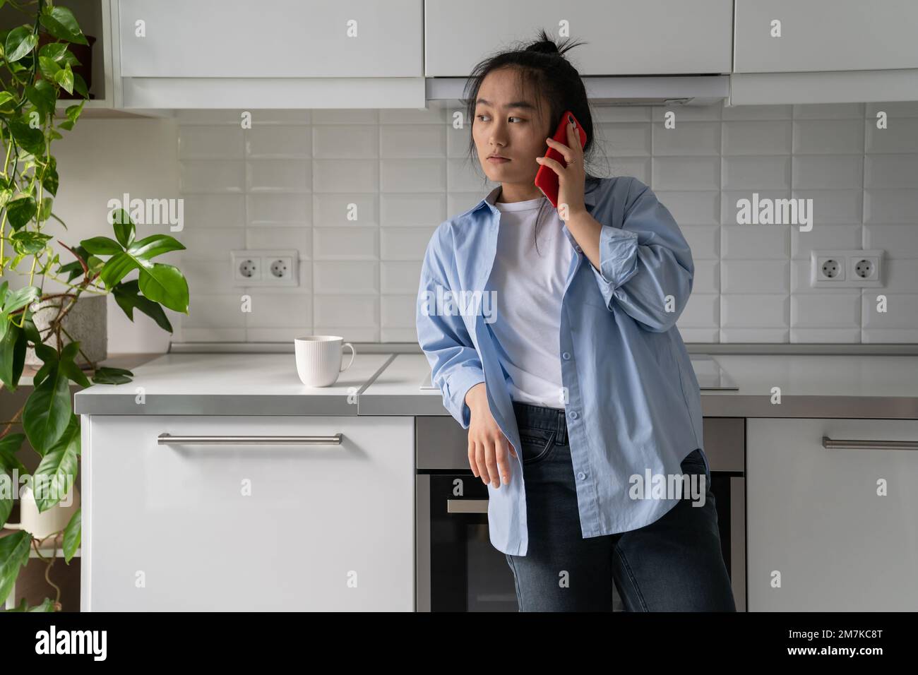 Tired pensive Asian woman standing in house at kitchen with coffee cup ...
