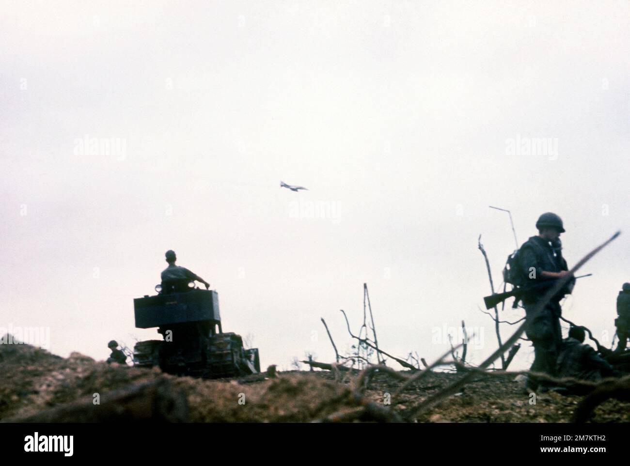 An F-4 Phantom II aircraft passes overhead as a bulldozer clears ...
