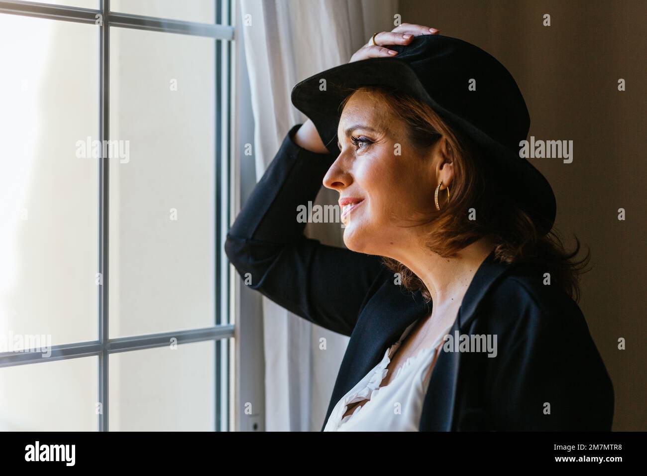 Woman holding hat on head while looking out window standing indoor at ...