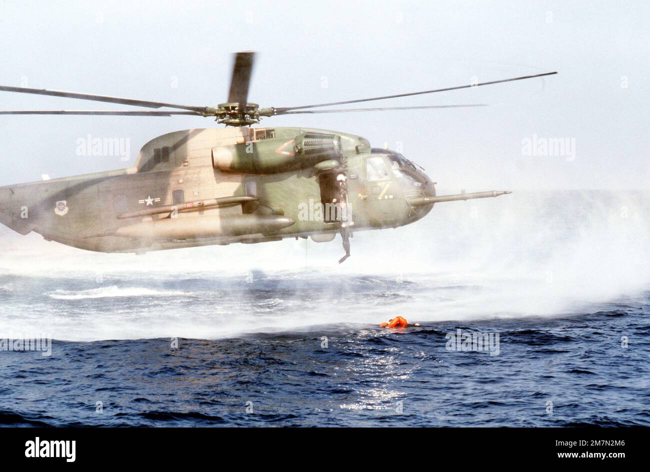 An HH-53 helicopter hovers low over the water as a pararescueman of the ...