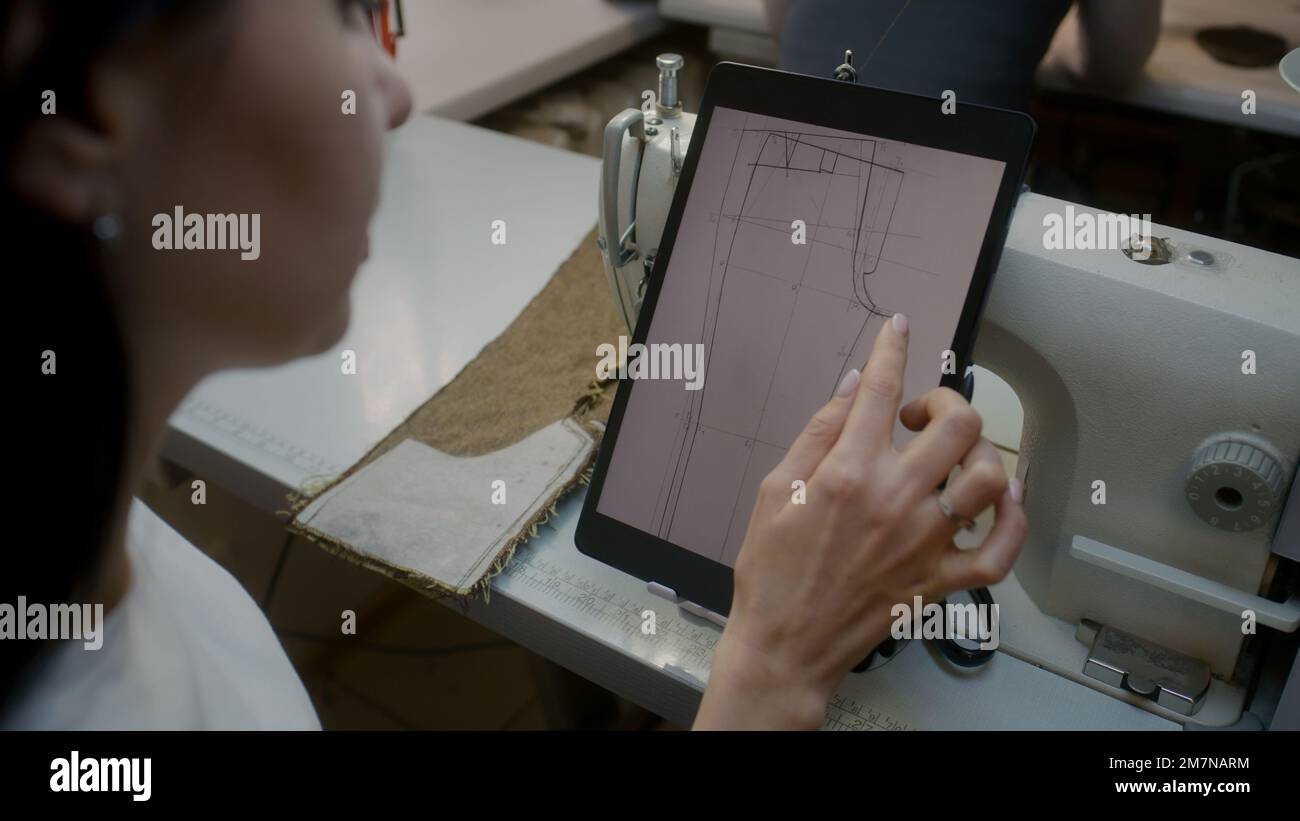 Female tailor sits at sewing machine with digital tablet and zooming ...