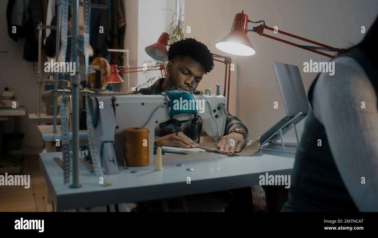 African American male seamstress sits at sewing machine and tailoring ...