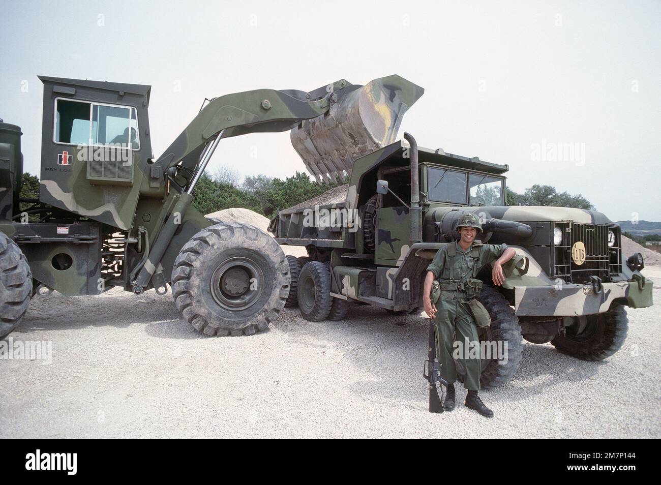 A US Army sentry guards a wheeled loader and an M51 dump truck. Country ...