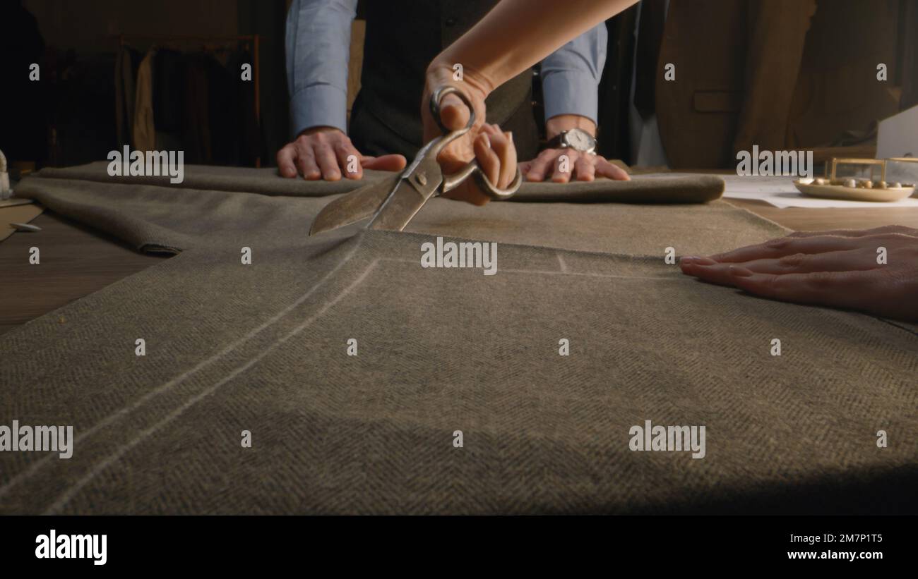 Close-up shot of female tailor cutting fabric according to sketch using scissors for sewing ...