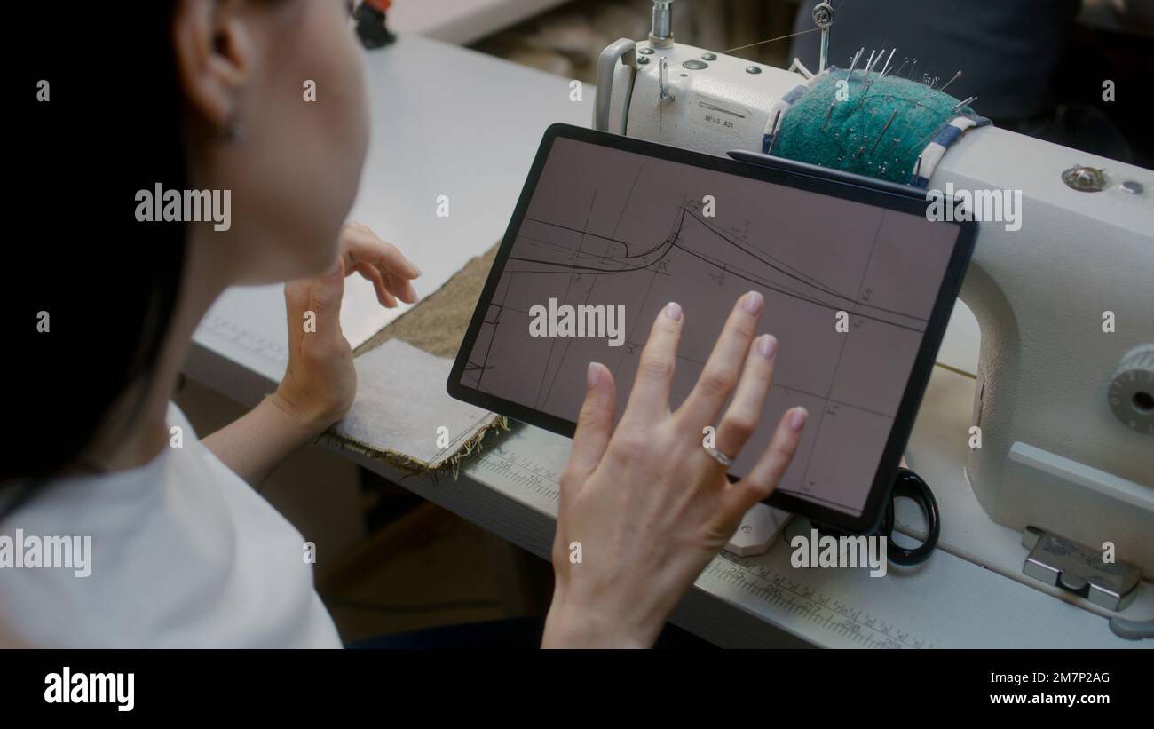 Female tailor sits at sewing machine with digital tablet and zooming ...