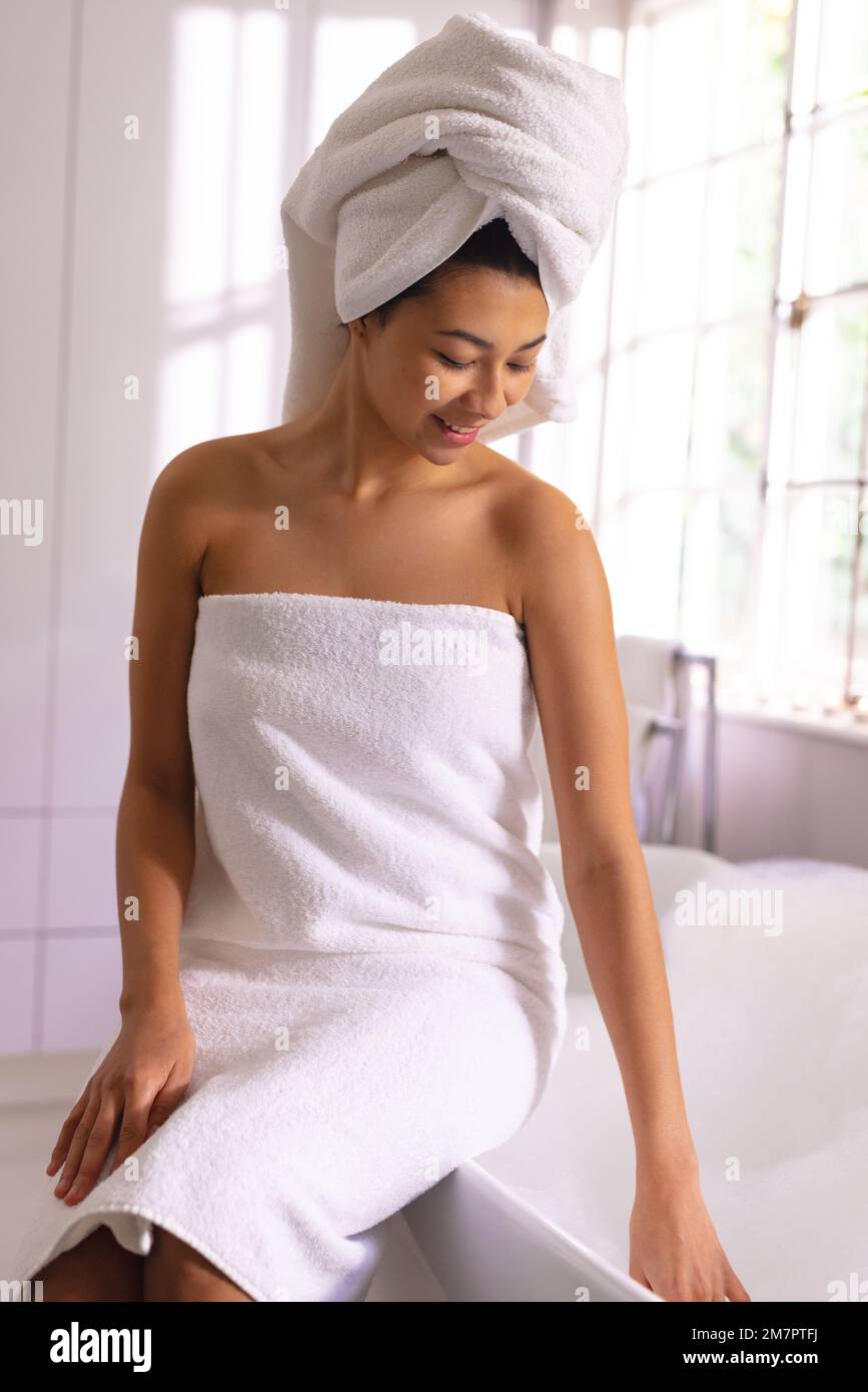 Vertical of happy biracial woman wearing towel sitting on the edge of ...