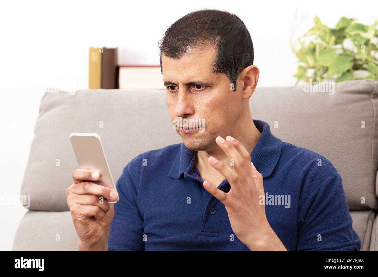 Angry man looking at smart phone sitting on a couch in the living room ...