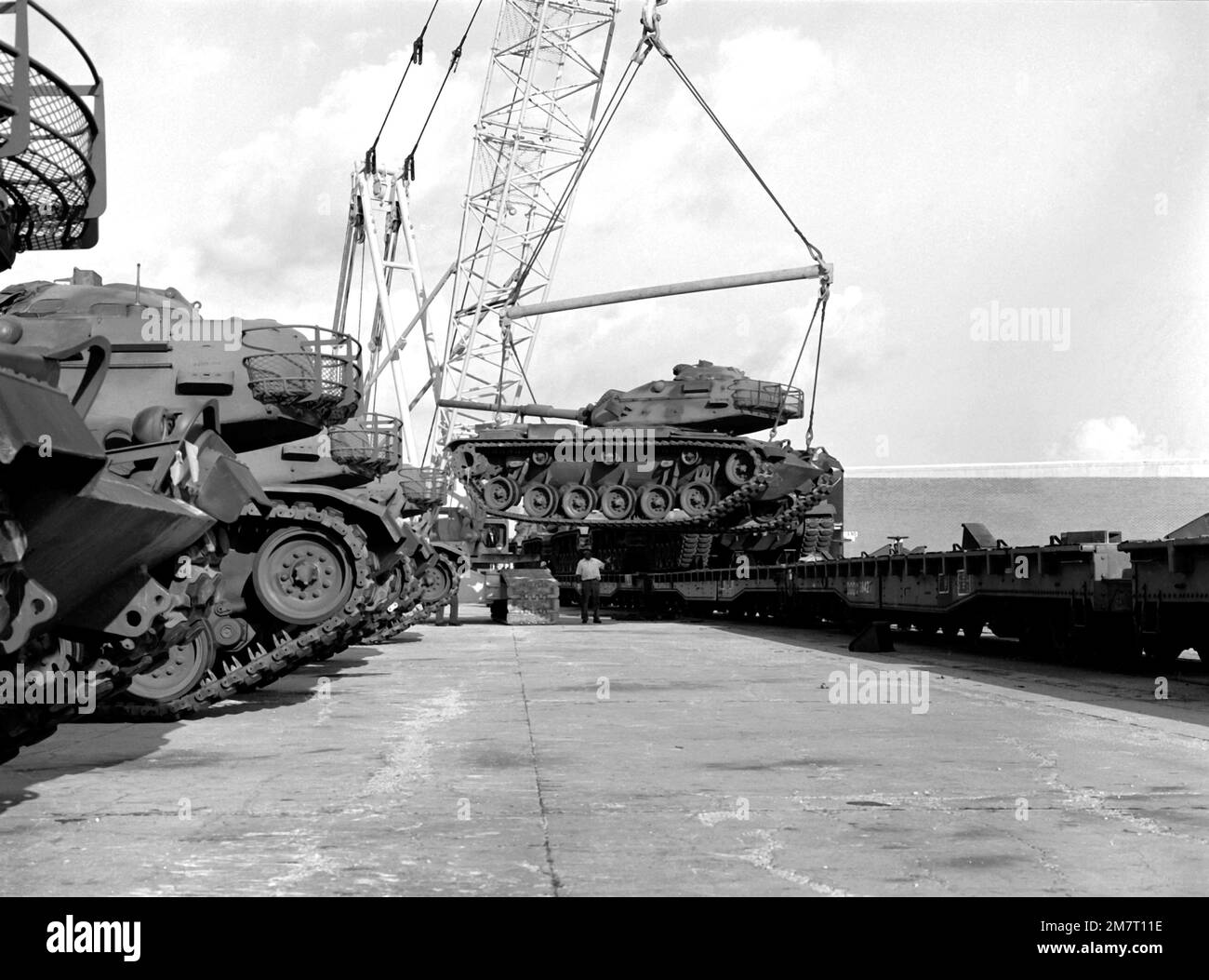 Base workmen use a huge crane to load M-60A1 tanks aboard railroad flat ...