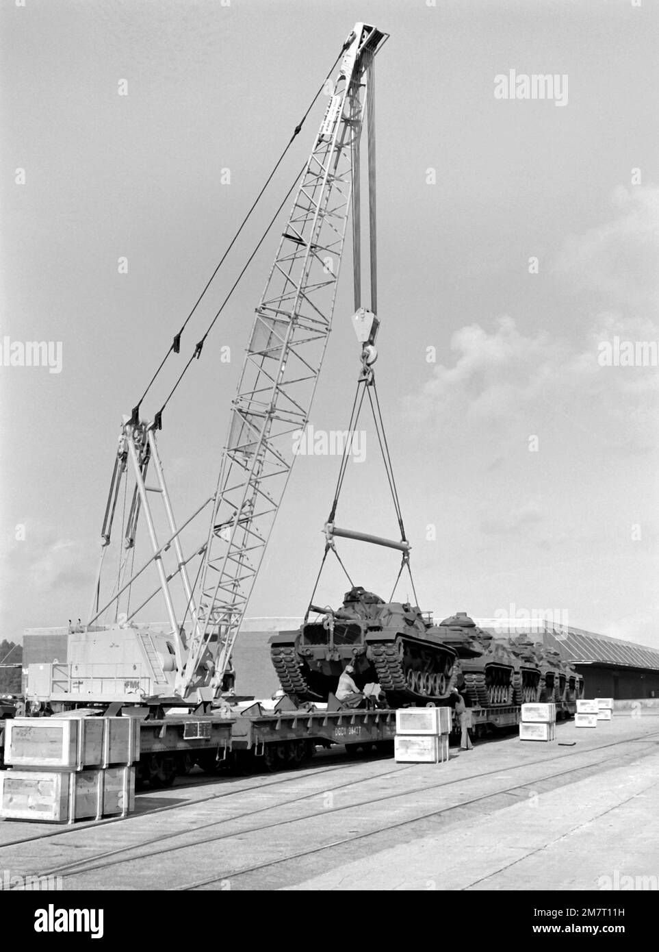 Base workmen use a huge crane to load M-60A1 tanks aboard railroad flat ...