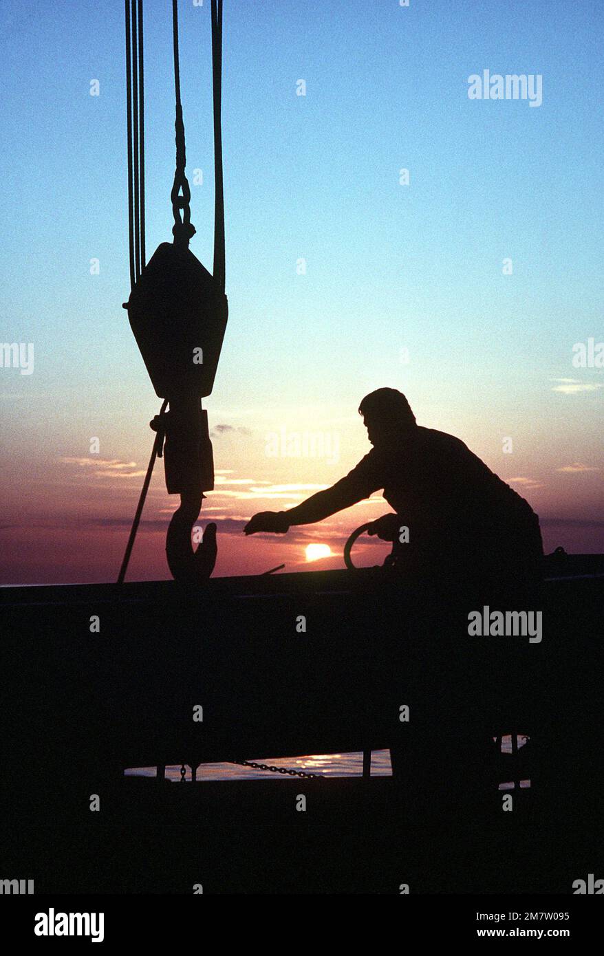 A crewman lowers a hook to load supplies aboard the surveying ship USNS ...