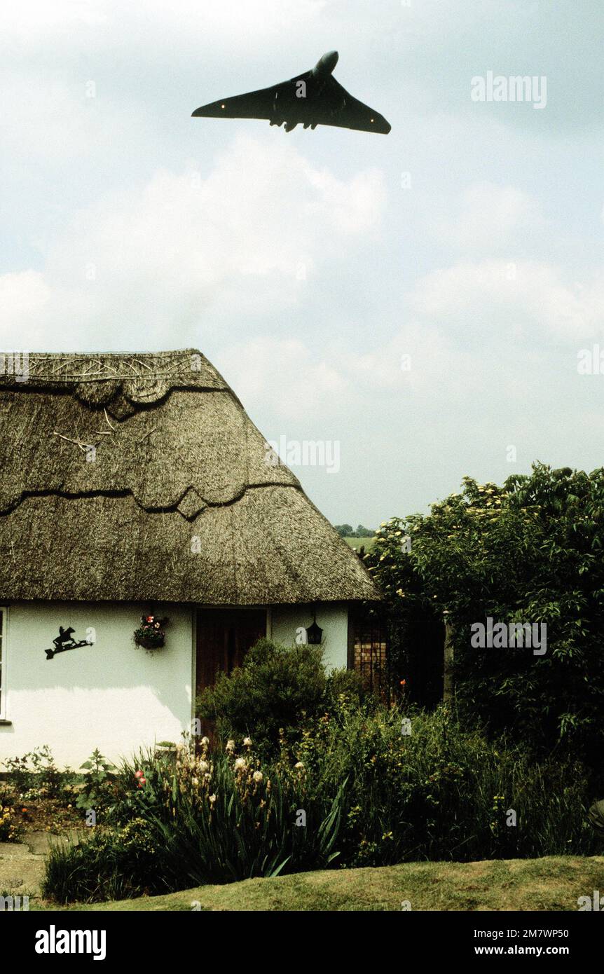 A view of a British Vulcan aircraft flying over a thatched roof house ...
