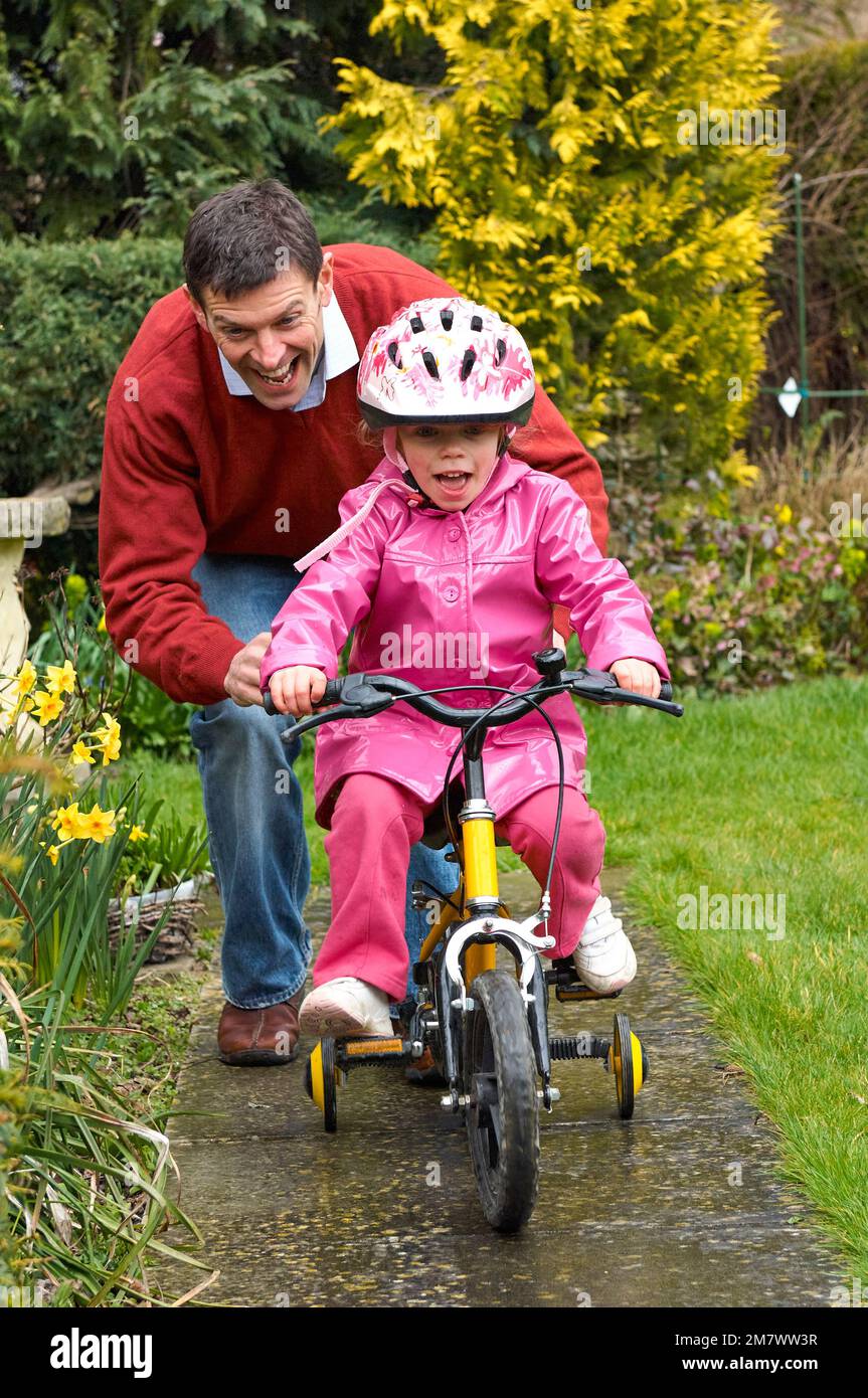 A man helping a young girl to ride her yellow bike with stabilisers on ...