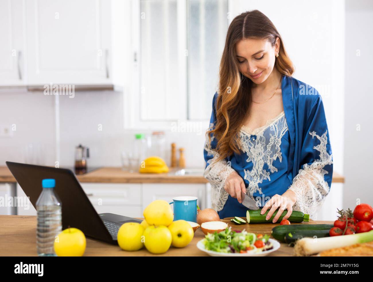 Housewife in housecoat reads recipe in laptop and prepares a salad in ...