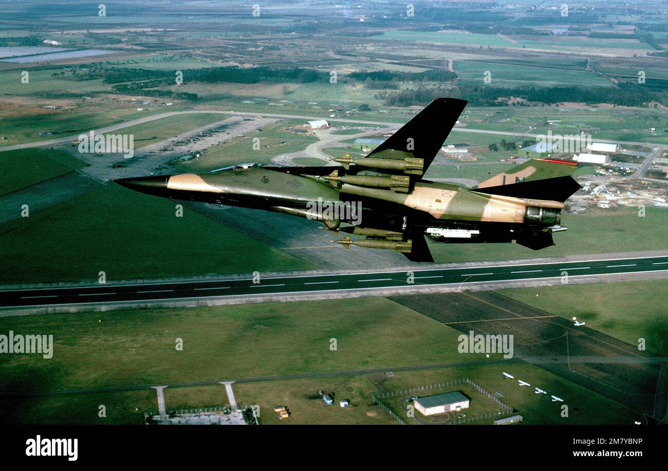 An air-to-air left underside view of an F-111 aircraft, armed with four bombs on its wing pylons ...