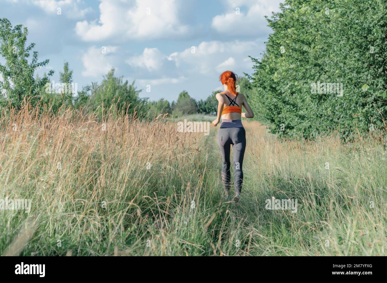 A young female runner runs along the road overgrown with grass. Rear ...