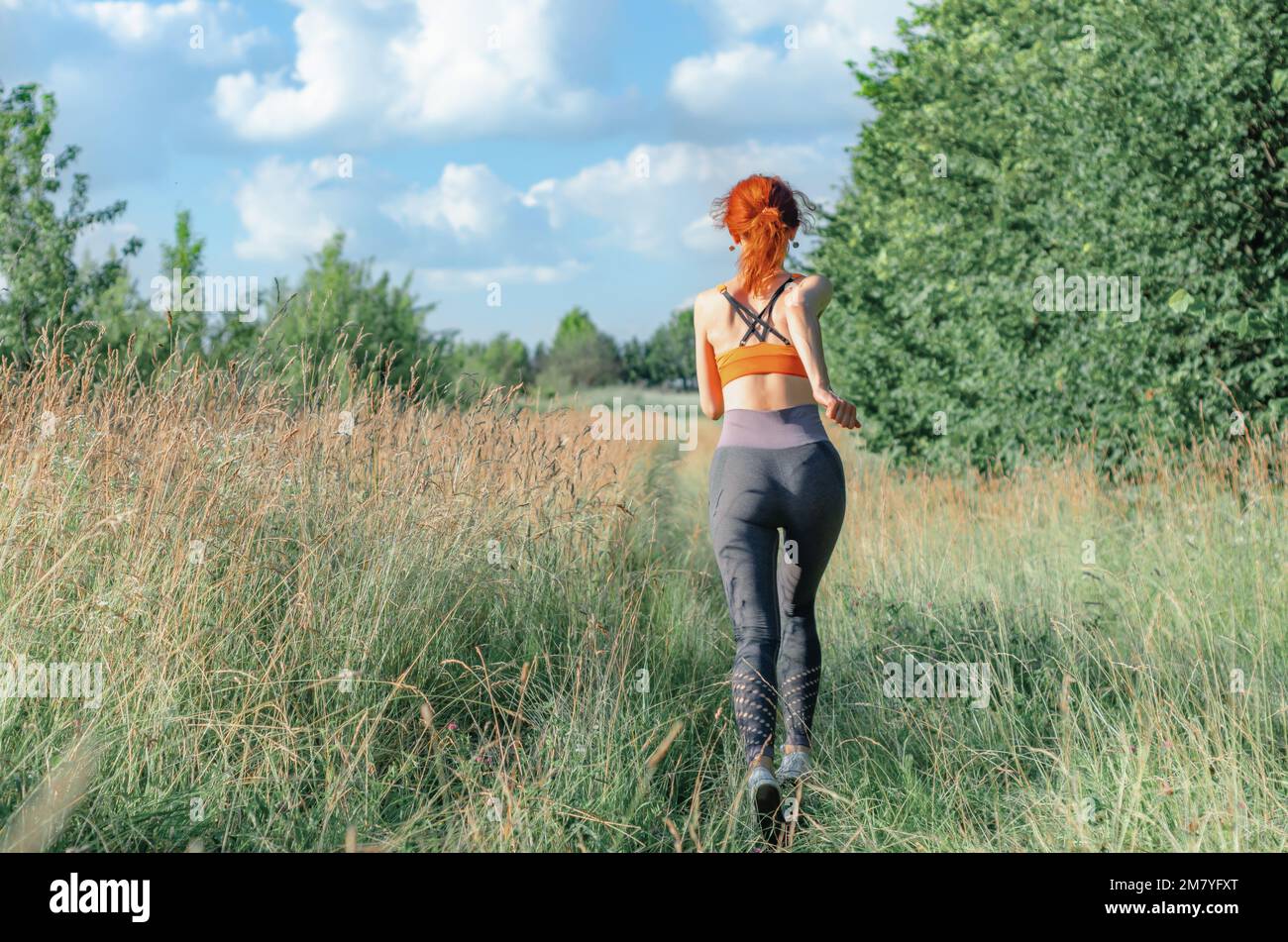 A young female runner runs along the road overgrown with grass. Rear ...