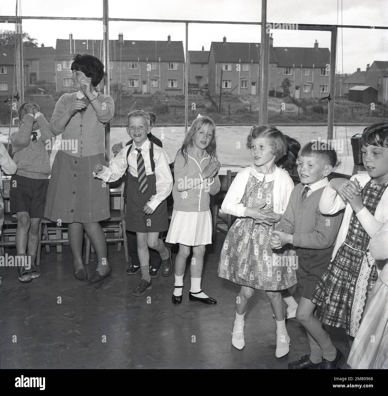 1960s historical, excited boys and girls in a pimary school classroom ...
