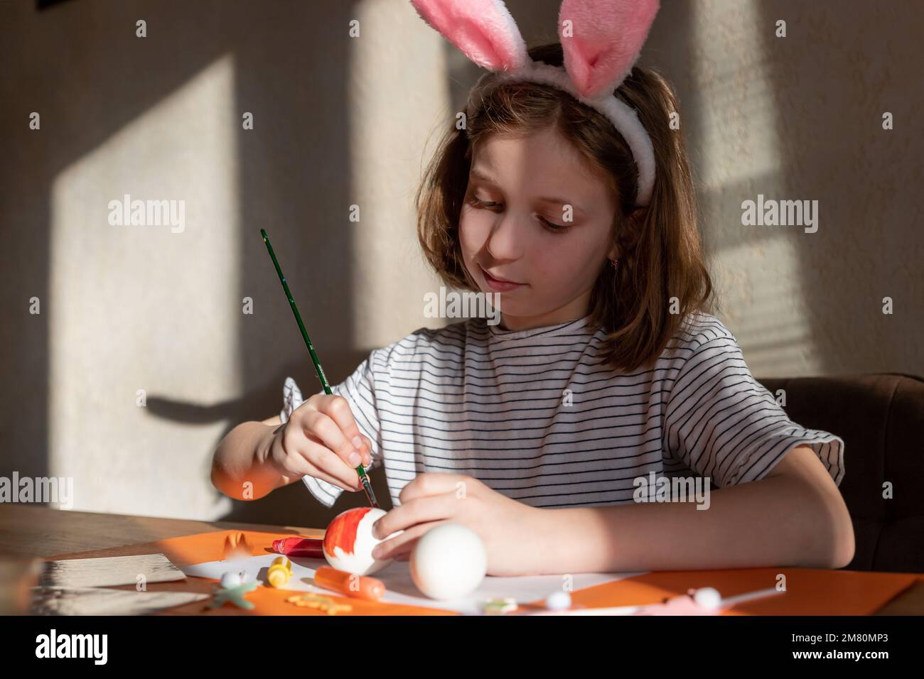 Cute little girl in bunny ears headband painting Easter eggs at table ...