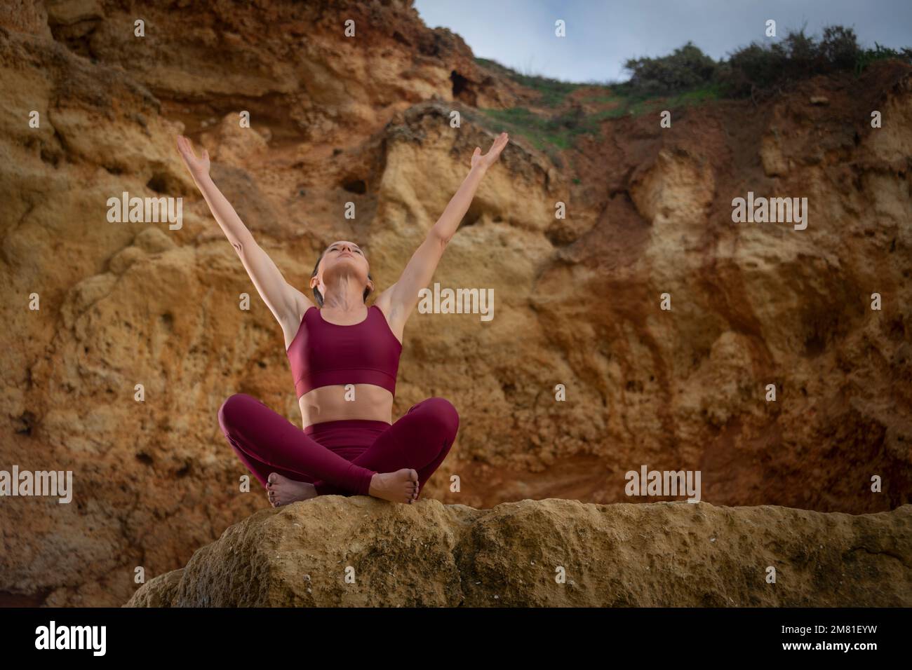 Woman sitting on rocks in lotus position with arms raised, yoga ...