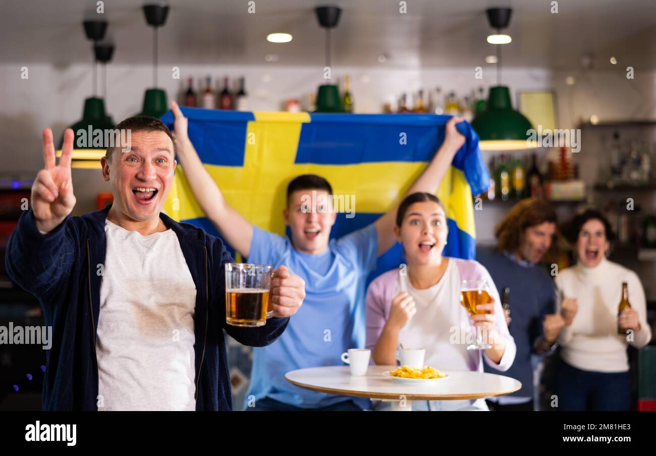 Company of excited young adult sports fans waving flag of Sweden and ...